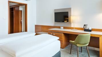 Quarto de hotel com cama, escrivaninha, cadeira, televisão e luminária.