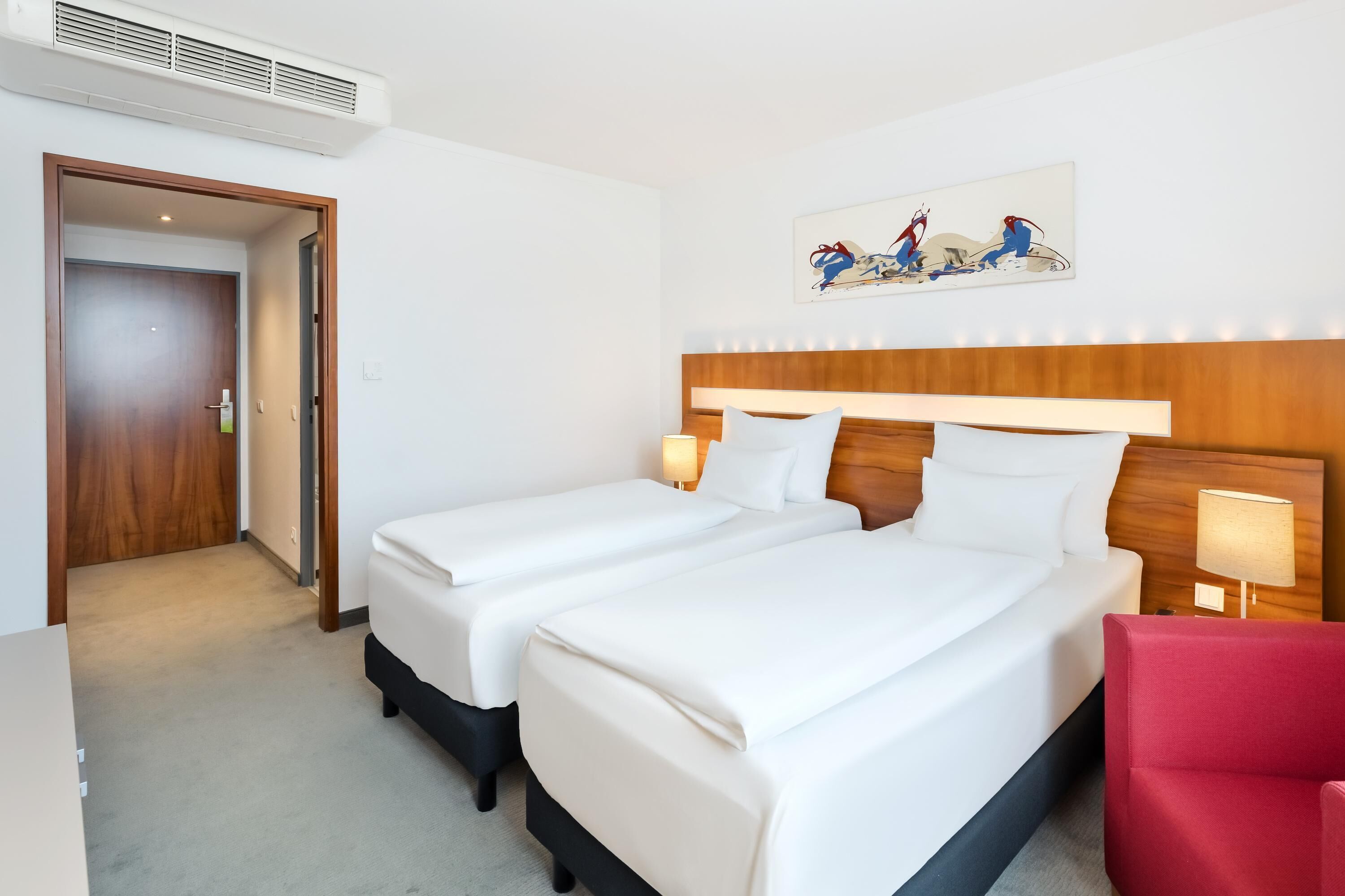 Quarto de hotel com duas camas, uma cadeira vermelha, uma cabeceira de madeira e uma obra de arte pendurada na parede.