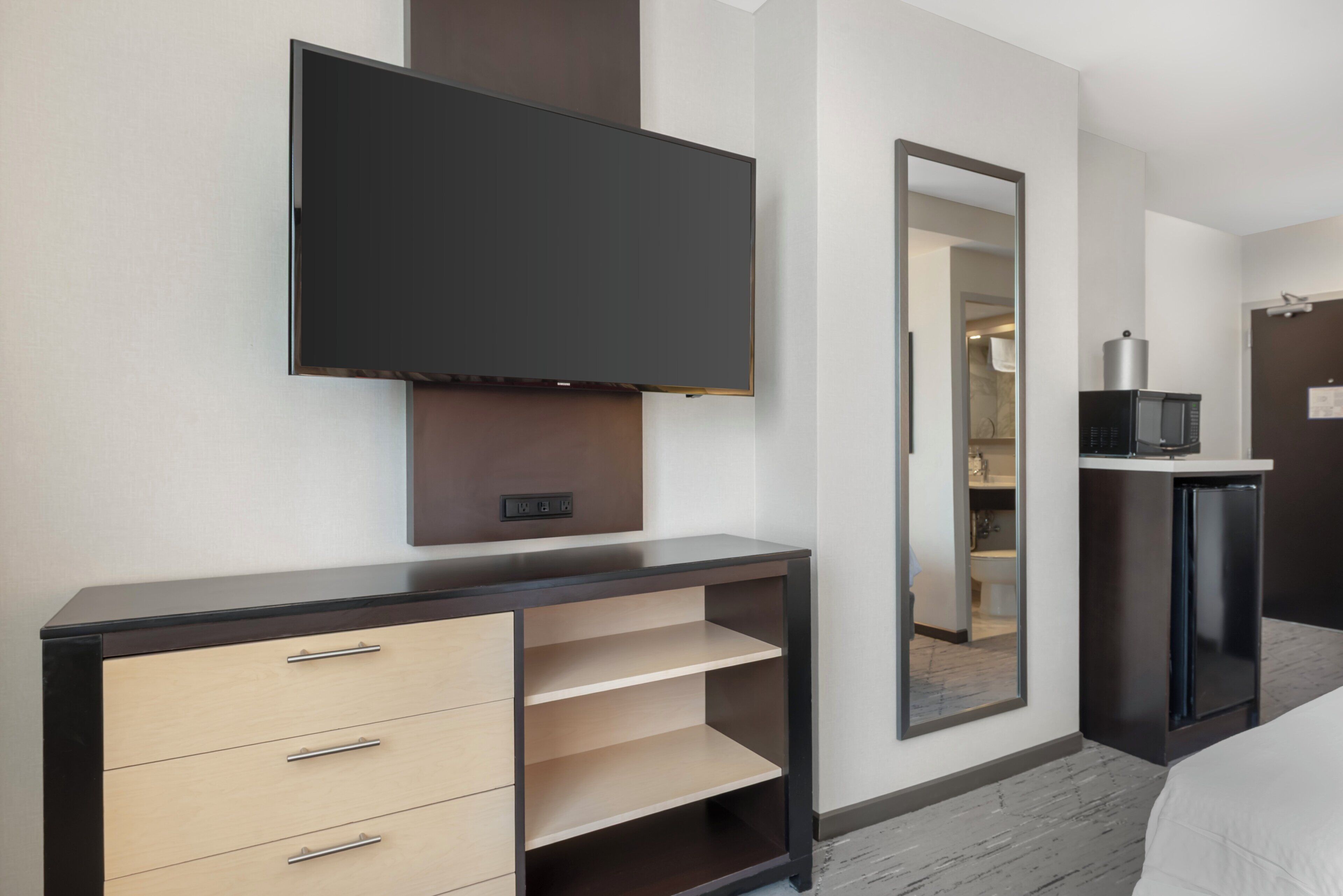 Quarto de hotel com televisão de tela plana na parede, espelho, um pequeno armário com micro-ondas e uma cama.