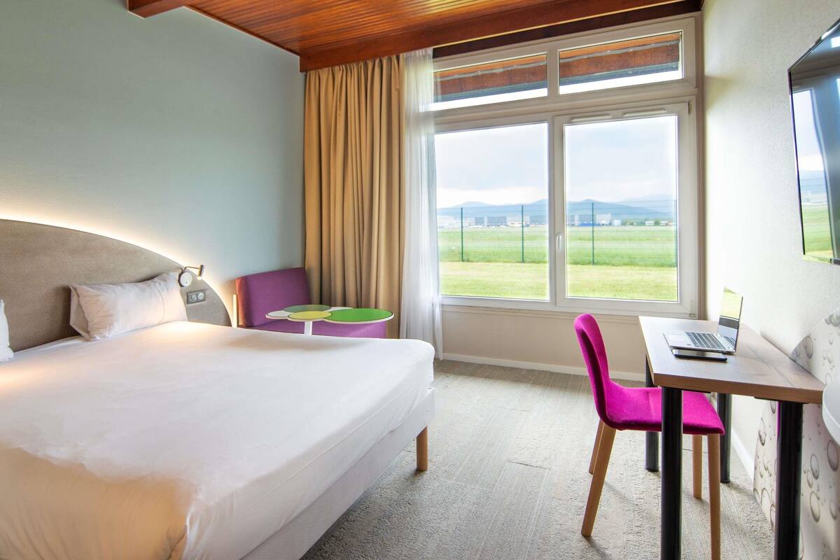 Quarto de hotel com uma cama, uma escrivaninha com um laptop, uma cadeira e vista para uma paisagem verde pela janela.