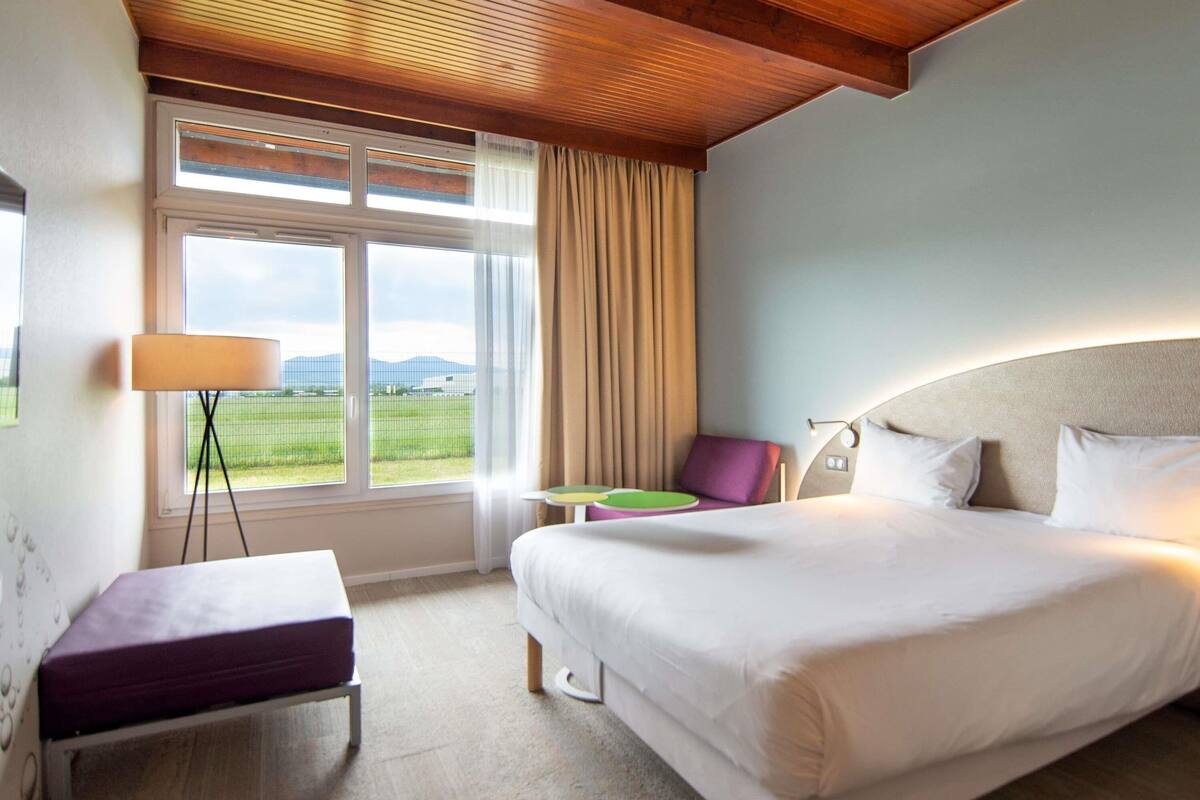 Quarto de hotel com cama, uma pequena mesa, uma cadeira, abajur e janela com cortinas.