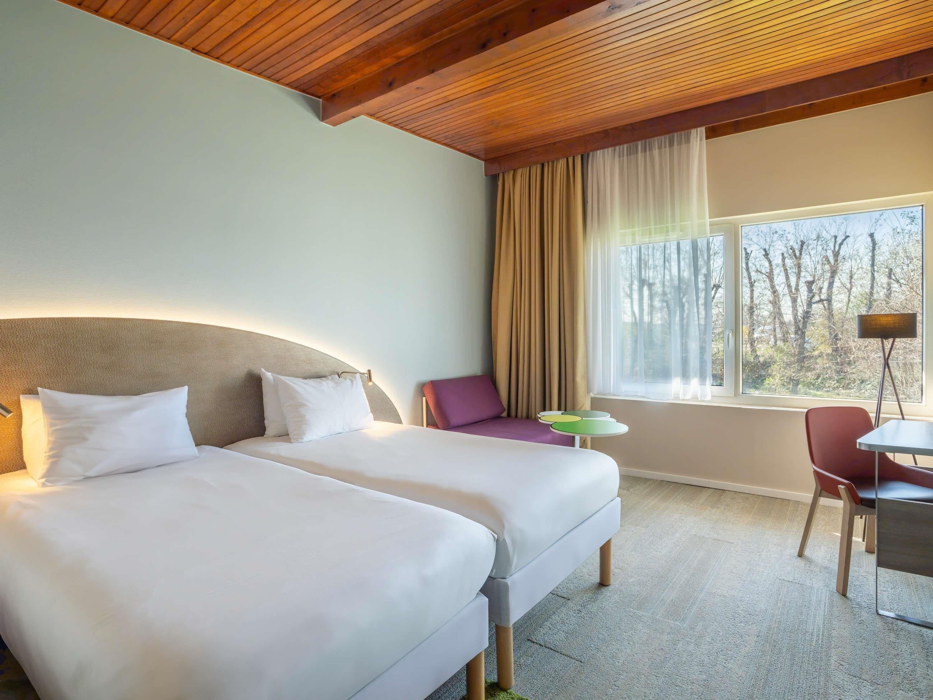 Quarto de hotel com duas camas, uma escrivaninha, uma cadeira e janela com vista para árvores.