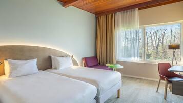 Quarto de hotel com duas camas, uma escrivaninha, uma cadeira e janela com vista para árvores.