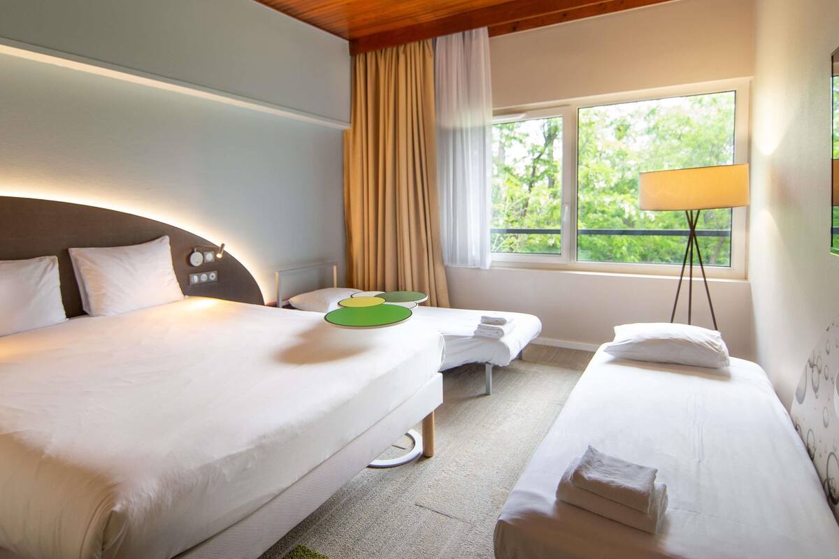 Quarto de hotel com duas camas, uma mesa de cabeceira, abajur e janela com vista para o verde.