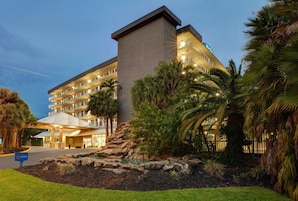 Um hotel de vários andares com design moderno, destacando uma entrada imponente com área coberta e um jardim paisagístico com palmeiras.