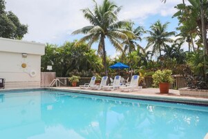 Uma piscina com espreguiçadeiras e guarda-sóis, cercada por palmeiras e plantas em vasos.