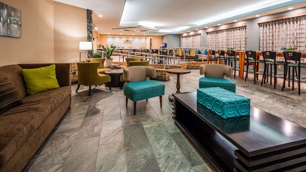 Um saguão de hotel com um sofá marrom, um banquinho azul-turquesa, poltronas amarelas e uma mesa de centro.