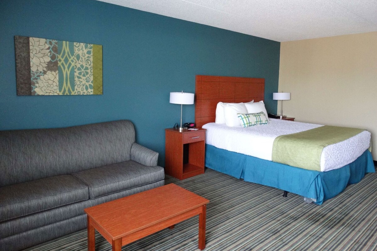 Quarto de hotel com cama, sofá, mesa de cabeceira, abajur e um quadro na parede.