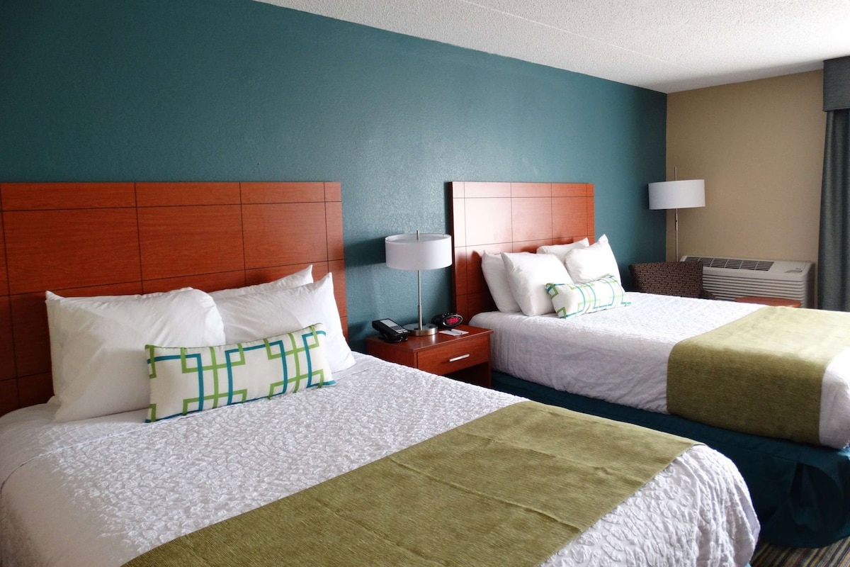 Quarto de hotel com duas camas, uma mesa de cabeceira com abajur, telefone e aparelho de ar condicionado.