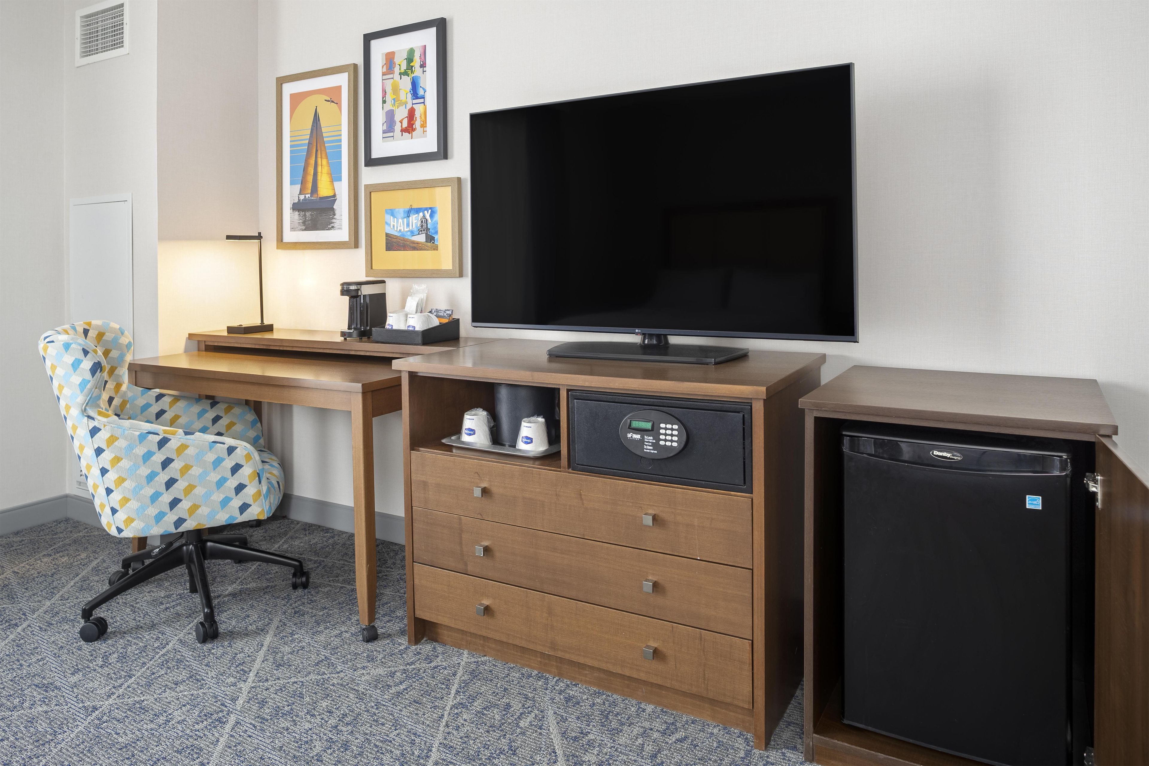 Quarto de hotel com escrivaninha, cadeira, televisão e mini-geladeira.