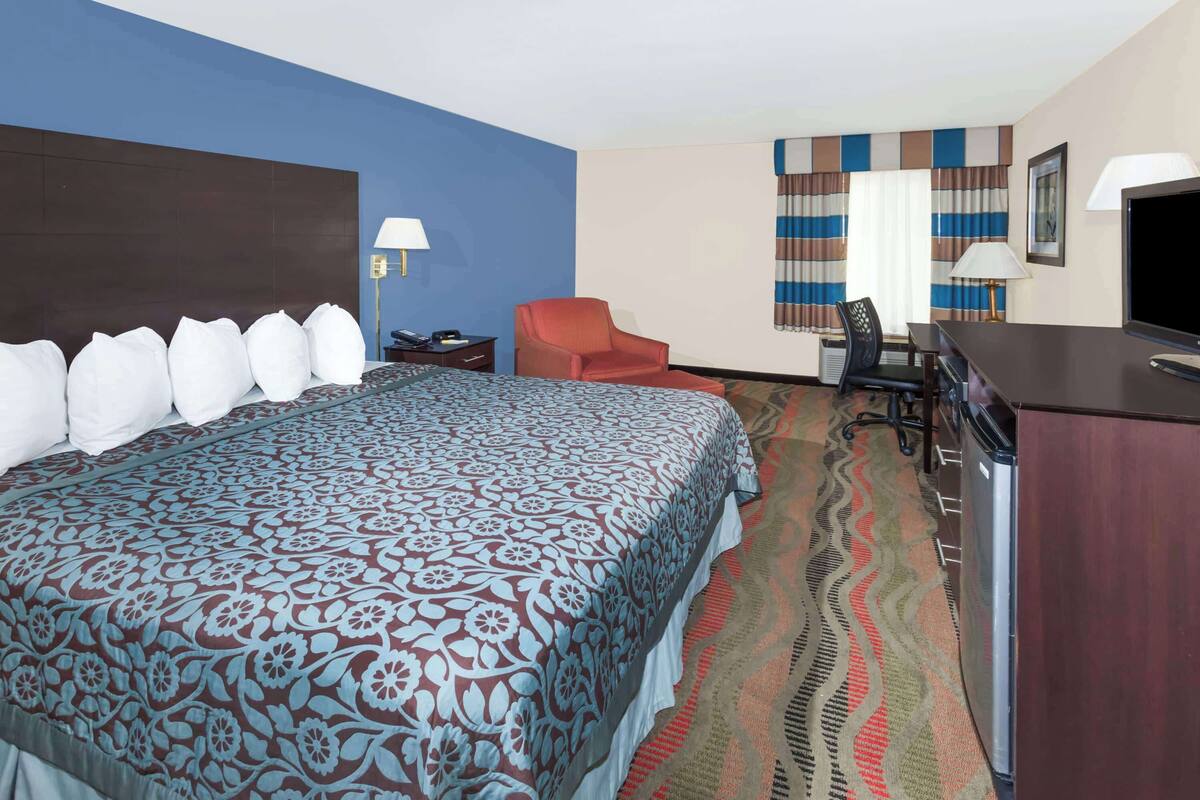 Quarto de hotel com cama grande, escrivaninha, cadeira, televisão e janela com cortinas.