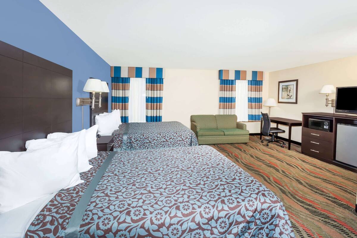 Quarto de hotel com duas camas, uma escrivaninha, uma cadeira, uma televisão e uma luminária de mesa.