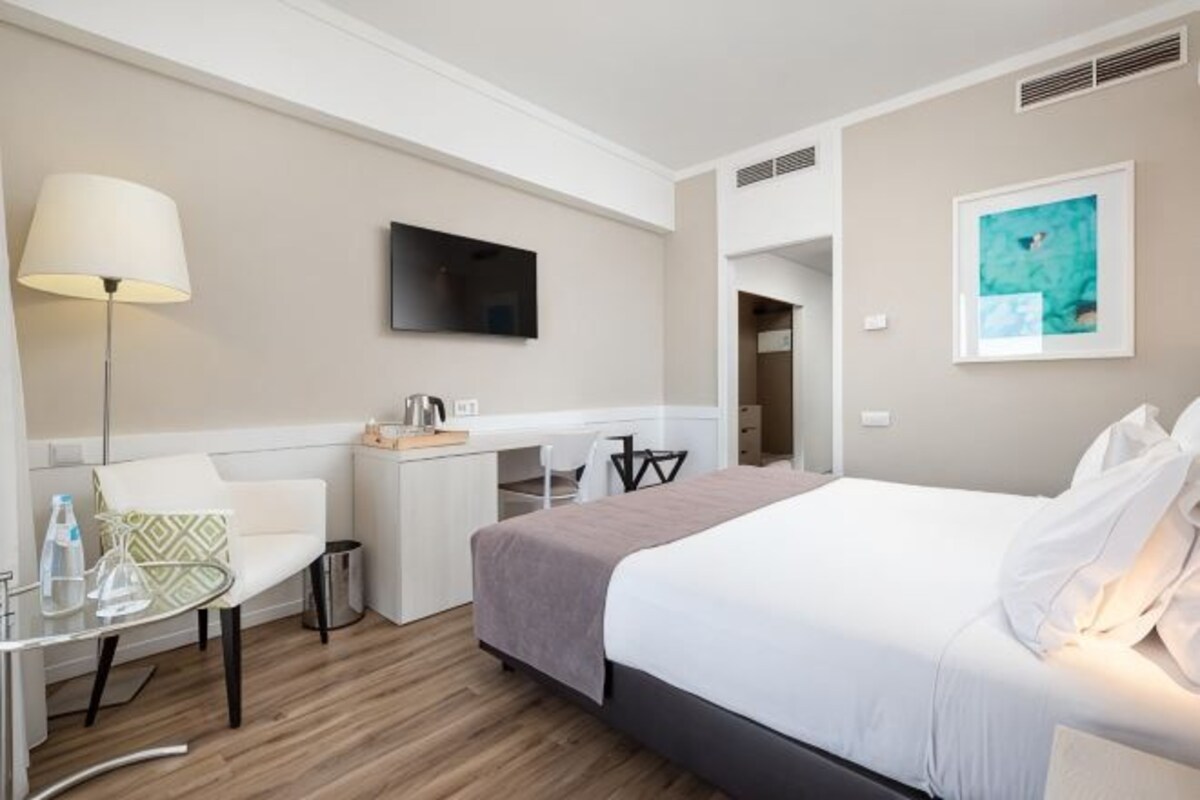 Quarto de hotel com cama, escrivaninha, cadeira, luminária, televisão e obras de arte na parede.