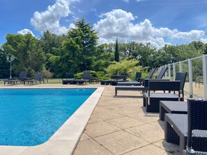 Uma piscina com espreguiçadeiras e mesas, cercada por árvores e um céu claro.