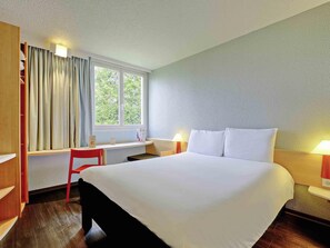 Quarto de hotel com cama, escrivaninha, cadeira e janela com cortinas.
