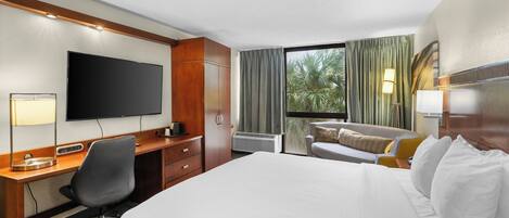 Quarto de hotel com uma cama grande, uma escrivaninha com cadeira, televisão de tela plana, janela com cortinas e vista para palmeiras.
