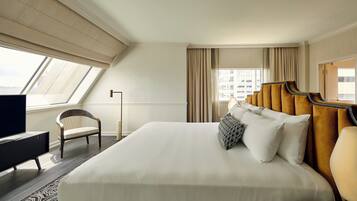 Quarto de hotel com uma cama grande, televisão, poltrona e vista da cidade pela janela.