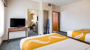 Quarto de hotel com duas camas, uma televisão sobre um console de madeira, uma escrivaninha com uma cadeira e um banheiro visível através de uma porta aberta.