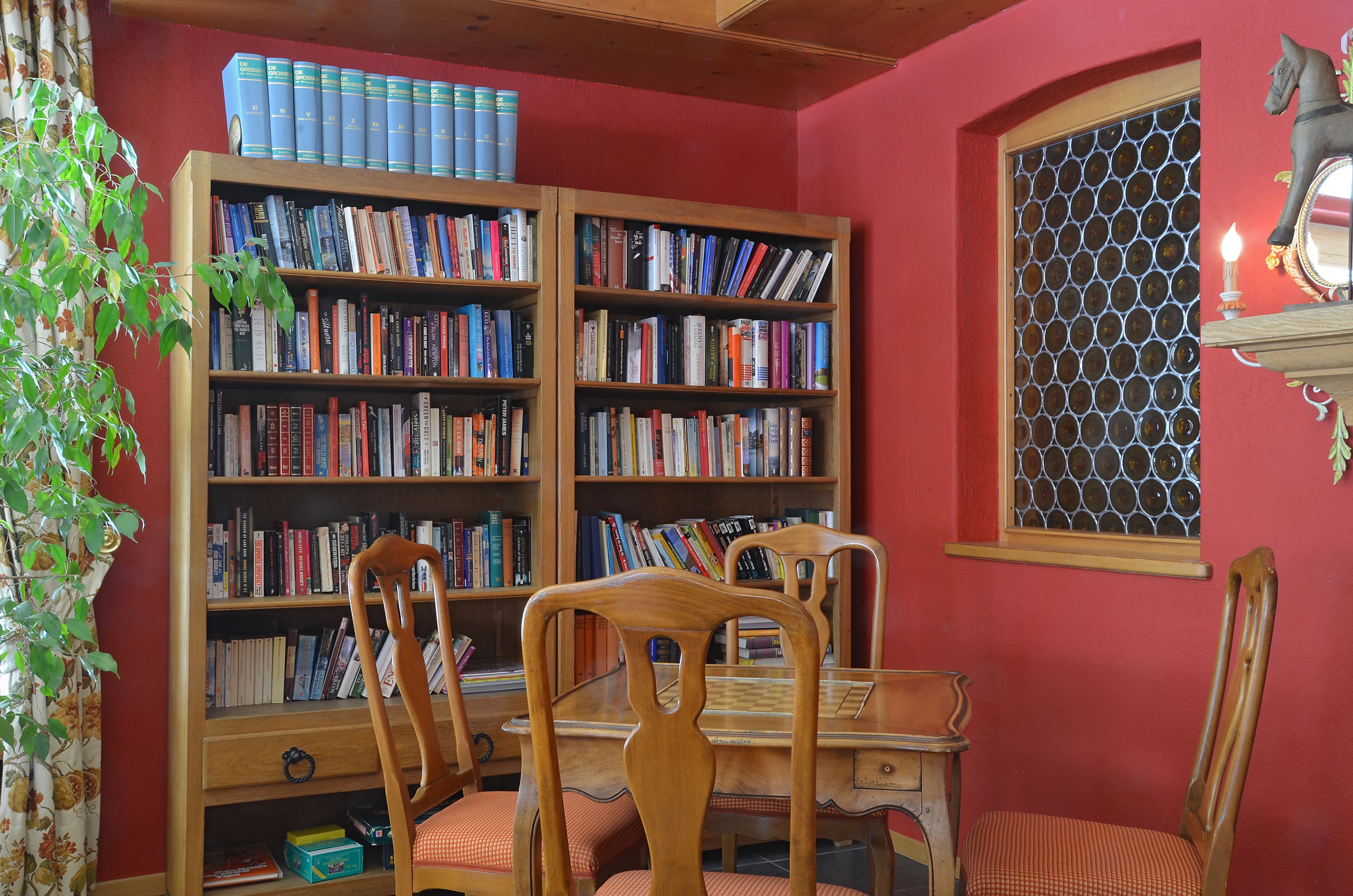 Uma biblioteca com estantes de madeira repletas de livros, uma mesa de madeira com cadeiras e uma janela com grade decorativa.