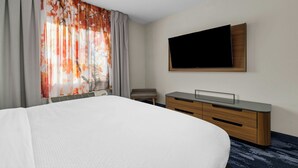 Quarto de hotel com uma cama grande, uma TV de tela plana na parede, um aparador de madeira e uma janela com cortinas finas.