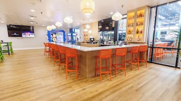 Uma área de bar moderna com piso de madeira, banquetas laranjas e uma televisão na parede.