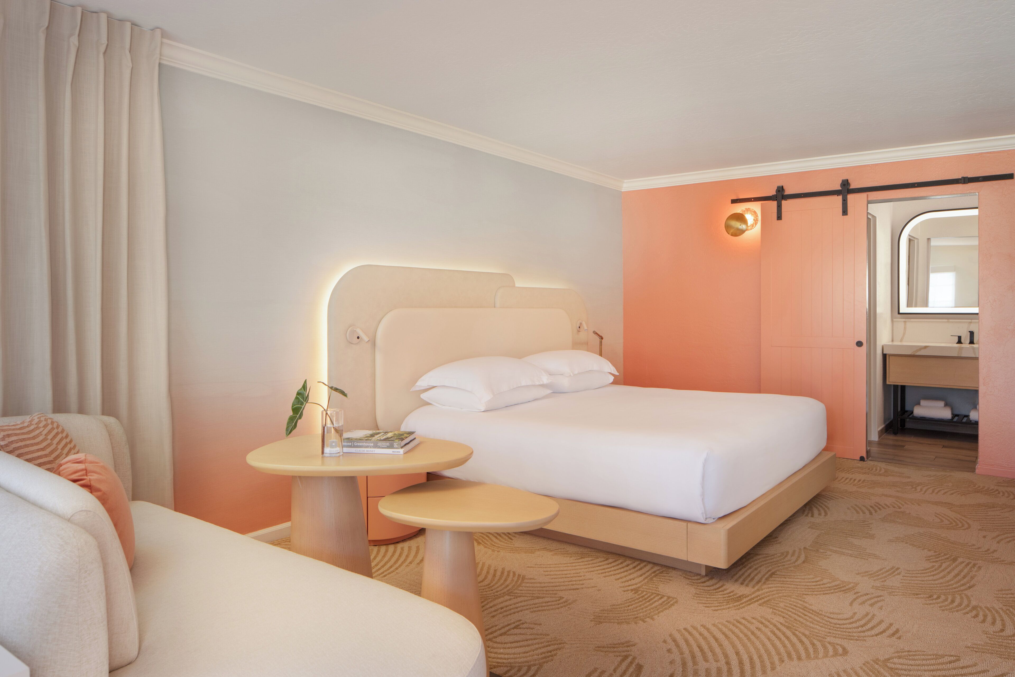 Quarto de hotel com cama, mesa de cabeceira com luminária, cadeira e pequena mesa com uma planta.