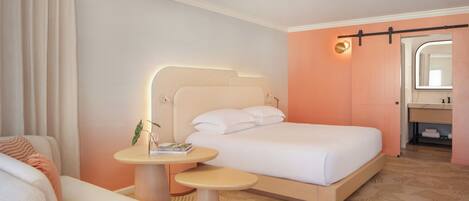 Quarto de hotel com cama, mesa de cabeceira com luminária, cadeira e pequena mesa com uma planta.