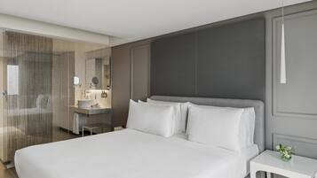 Quarto de hotel moderno com uma cama grande, uma mesa de cabeceira com um vaso de flores, um banheiro com espelho e pia, e uma porta de correr que dá para a área do chuveiro.