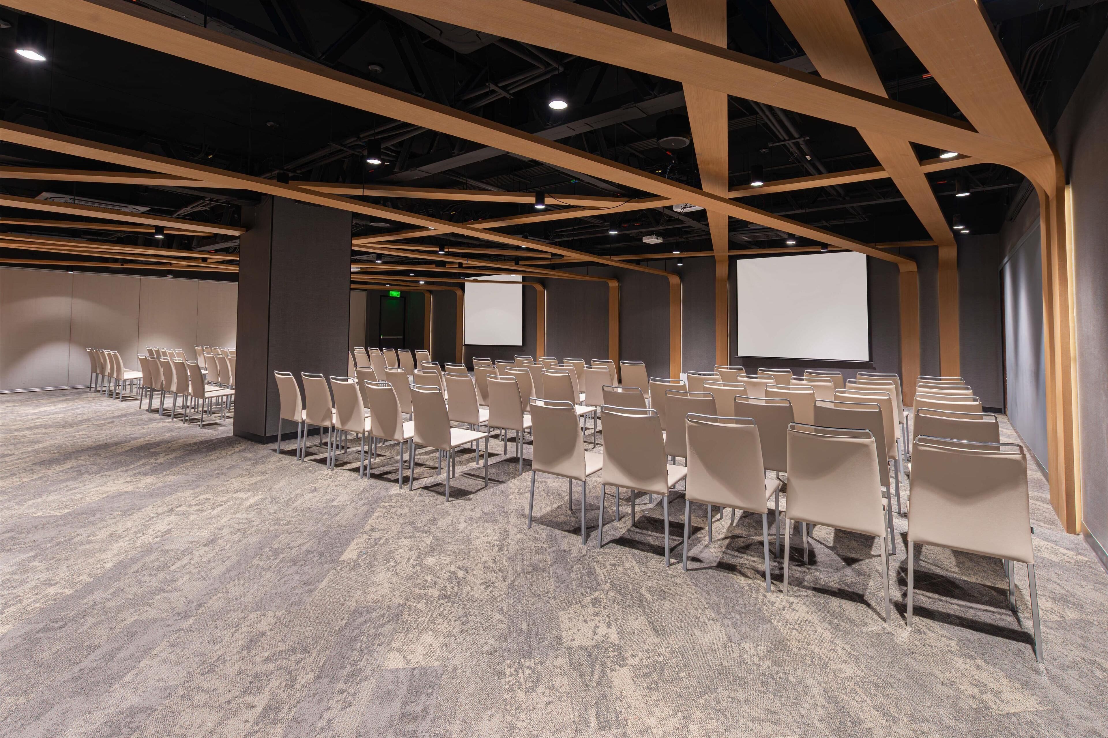 Uma sala de conferências com fileiras de cadeiras, uma tela grande e vigas de madeira no teto.