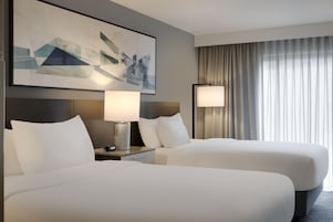 Quarto de hotel com duas camas, uma pintura abstrata grande, uma mesinha de cabeceira com abajur e uma janela com cortinas.