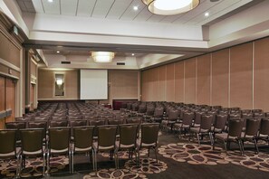 Sala de conferências com fileiras de cadeiras, uma tela de projetor e piso coberto de carpete.