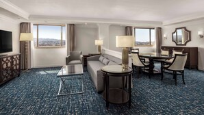 Quarto de hotel com sofá, mesa de centro, mesa de jantar com cadeiras, televisão e janelas grandes com cortinas.