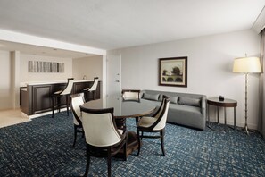 Quarto de hotel com mesa de jantar, cadeiras, sofá, luminária e um quadro na parede.