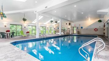 Piscina interior com água azul cristalina, rodeada por espreguiçadeiras e mesas, e grandes janelas que permitem a entrada de luz natural.