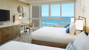 Quarto de hotel com duas camas, uma escrivaninha, uma cadeira, televisão e vista para o oceano.