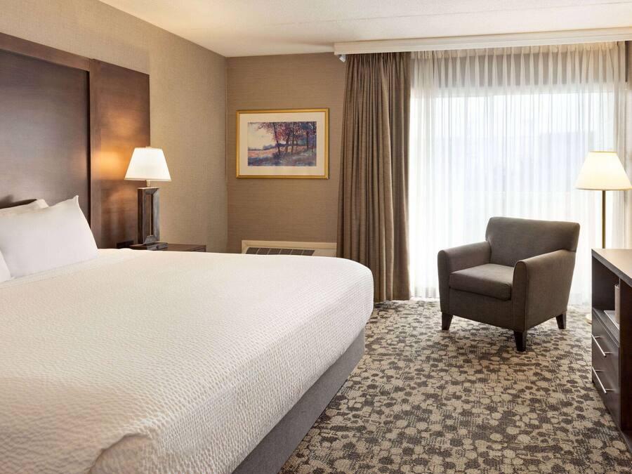 Quarto de hotel com uma cama grande, uma cadeira, uma escrivaninha com televisão e um quadro na parede.