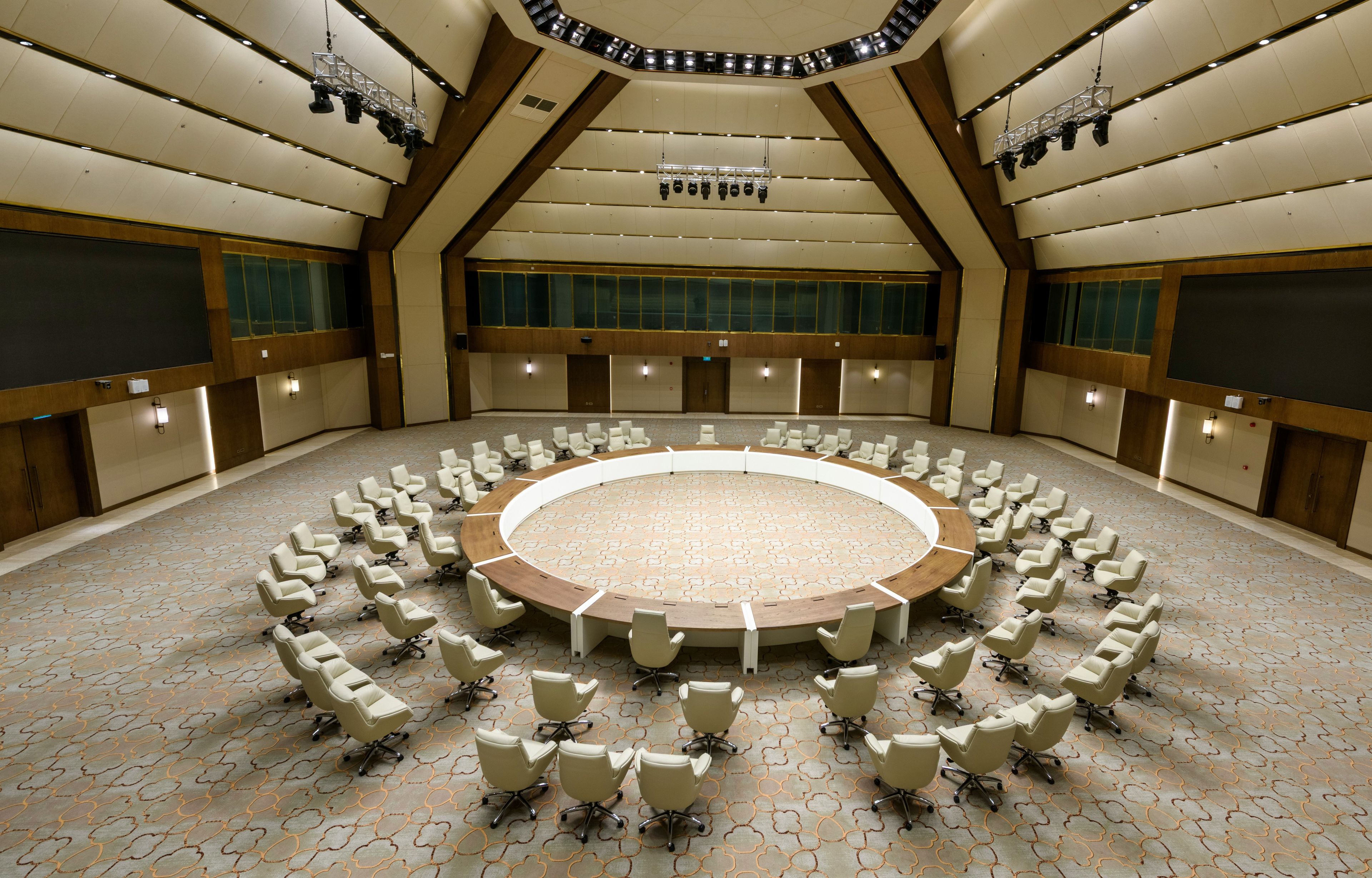 Sala de conferências com mesa redonda rodeada de cadeiras, tetos altos com luminárias e um tapete estampado.