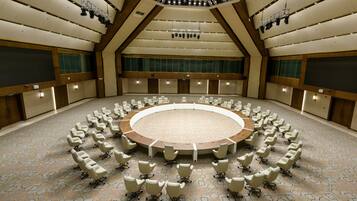 Sala de conferências com mesa redonda rodeada de cadeiras, tetos altos com luminárias e um tapete estampado.