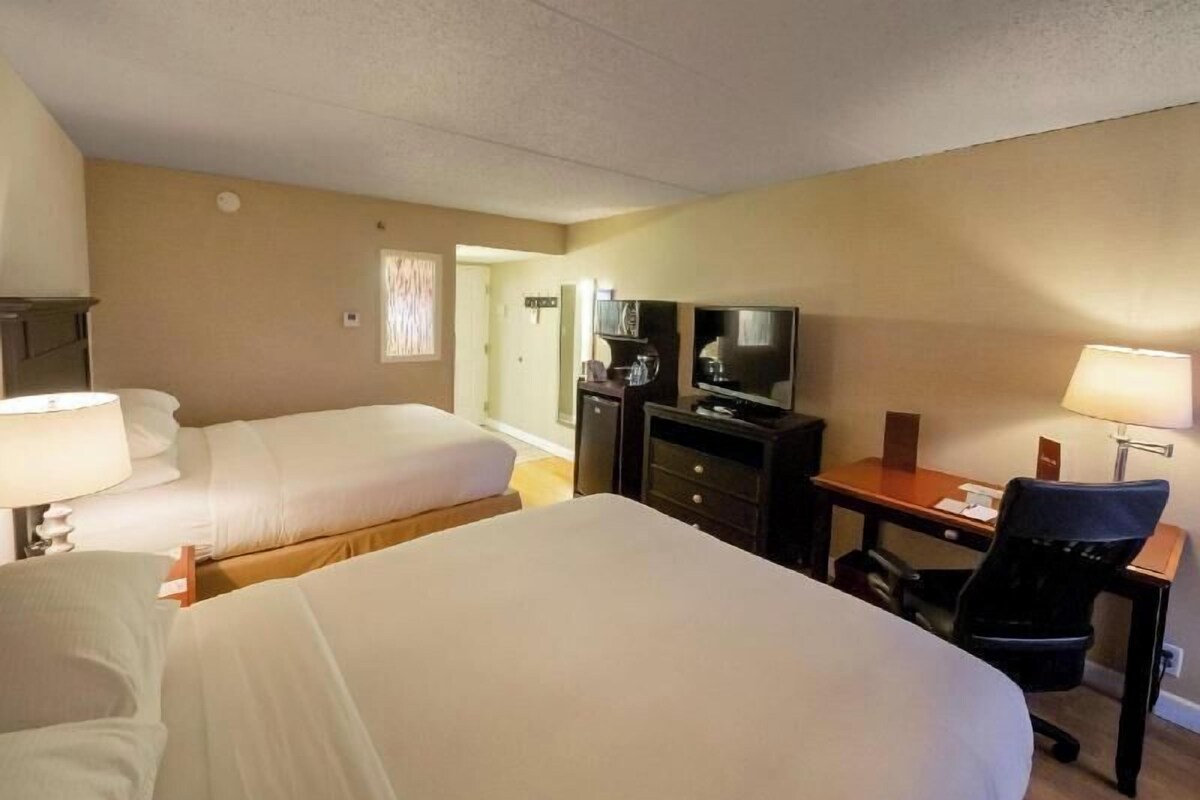 Quarto de hotel com duas camas, uma mesa de trabalho com cadeira, televisão e luminária.