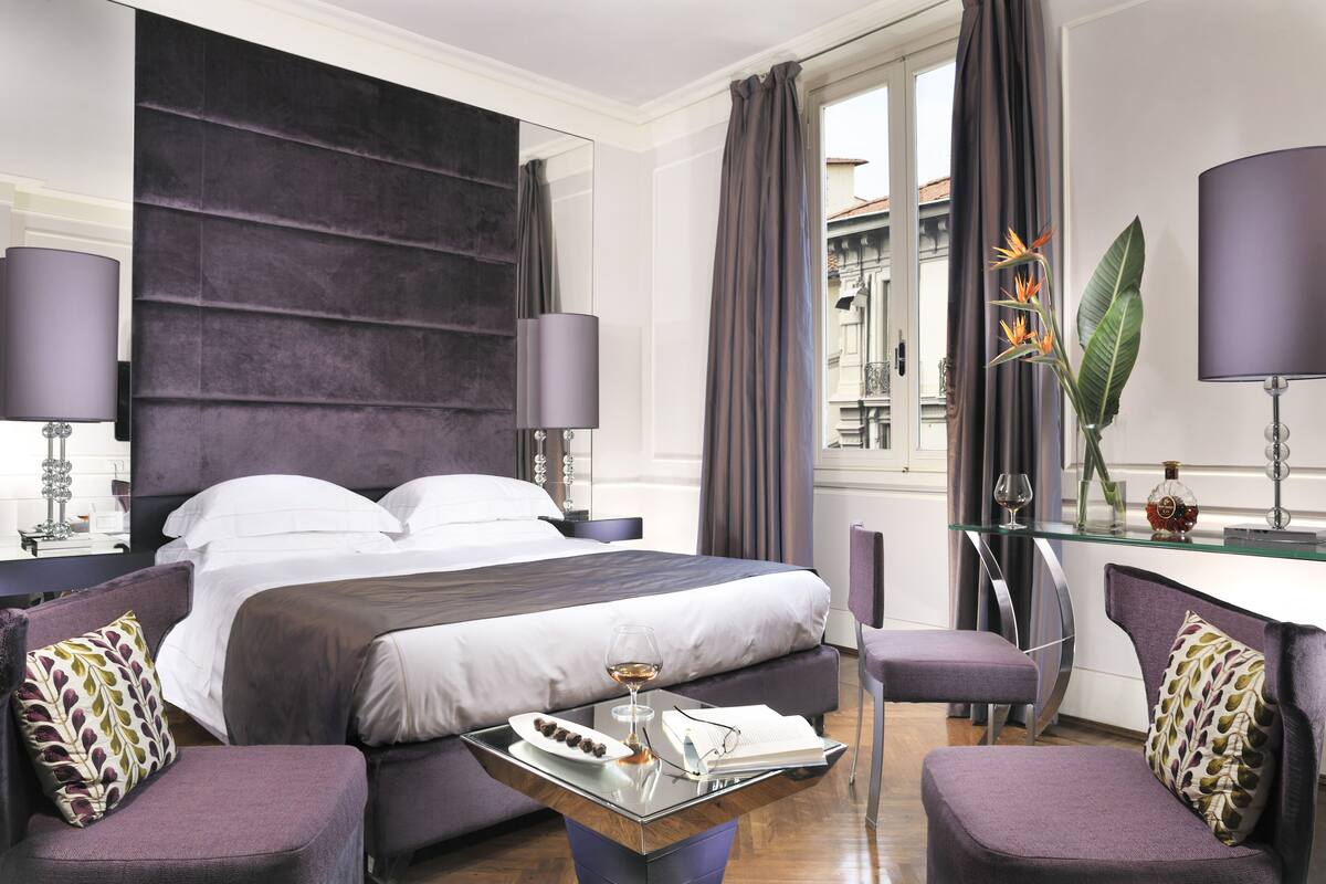 Quarto de hotel moderno com uma cama grande, uma mesa de vidro com uma taça de vinho e um livro, duas poltronas, uma escrivaninha com uma luminária e vista para a cidade através de grandes janelas.