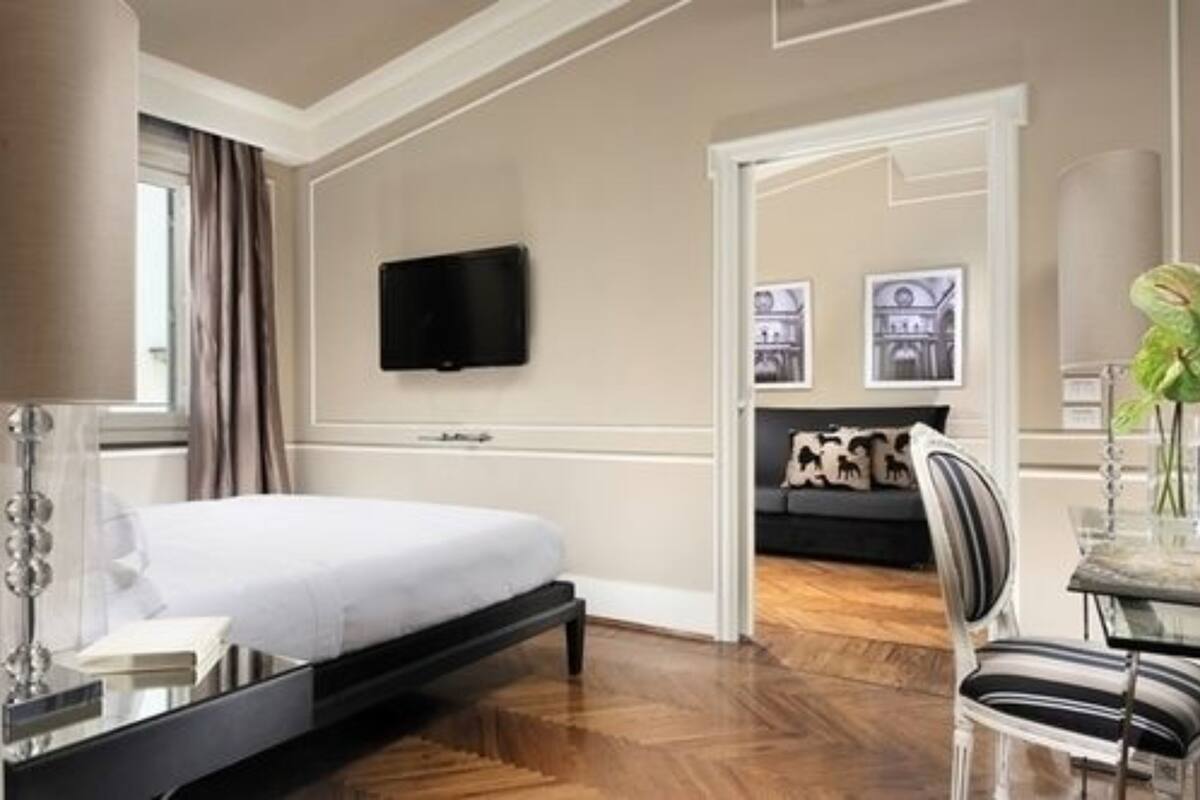 Quarto de hotel com cama, televisão na parede, área de jantar com mesa e cadeiras e vista para o corredor.
