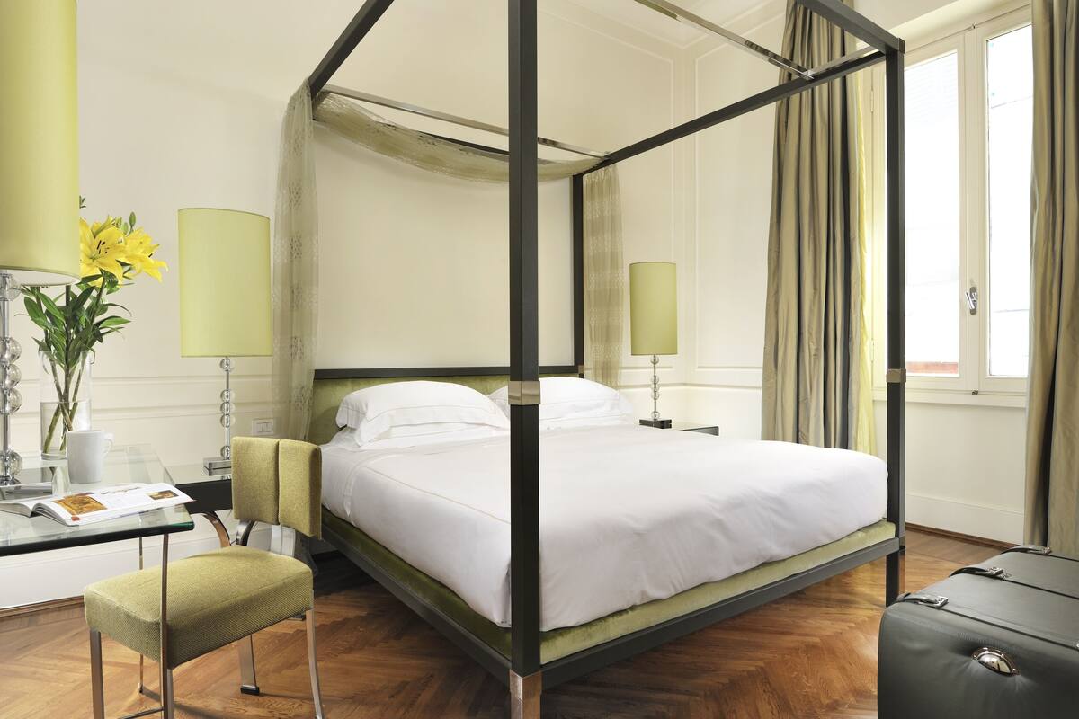 Uma cama de dossel com roupa de cama branca, uma mesinha de cabeceira com abajur, uma cadeira e uma mala em quarto de hotel.