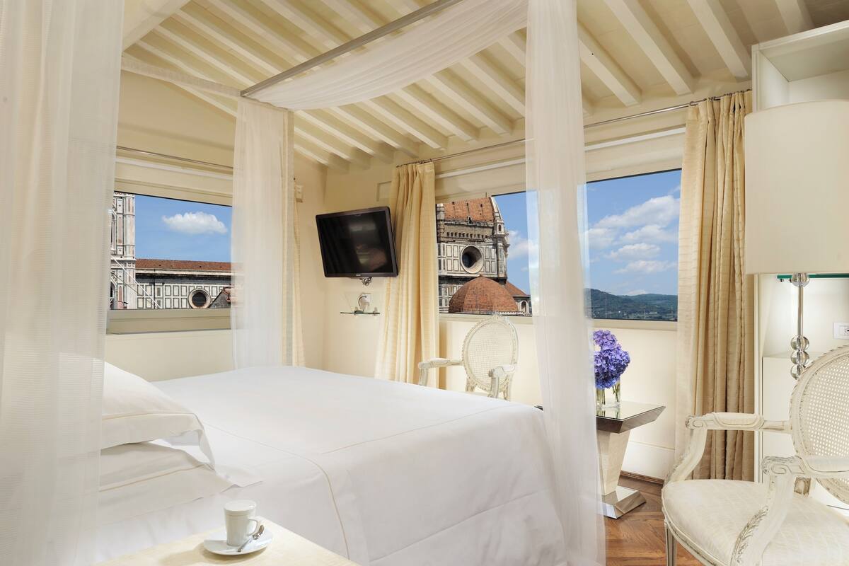 Quarto de hotel com cama, televisão, mesa com um vaso de flores e vista para um edifício histórico.