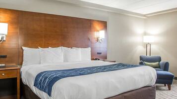 Quarto de hotel com uma cama grande, cabeceira de madeira, escrivaninha com abajur e uma cadeira.