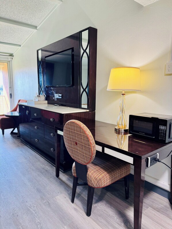 Quarto de hotel com escrivaninha, cadeira, televisão, luminária e micro-ondas.