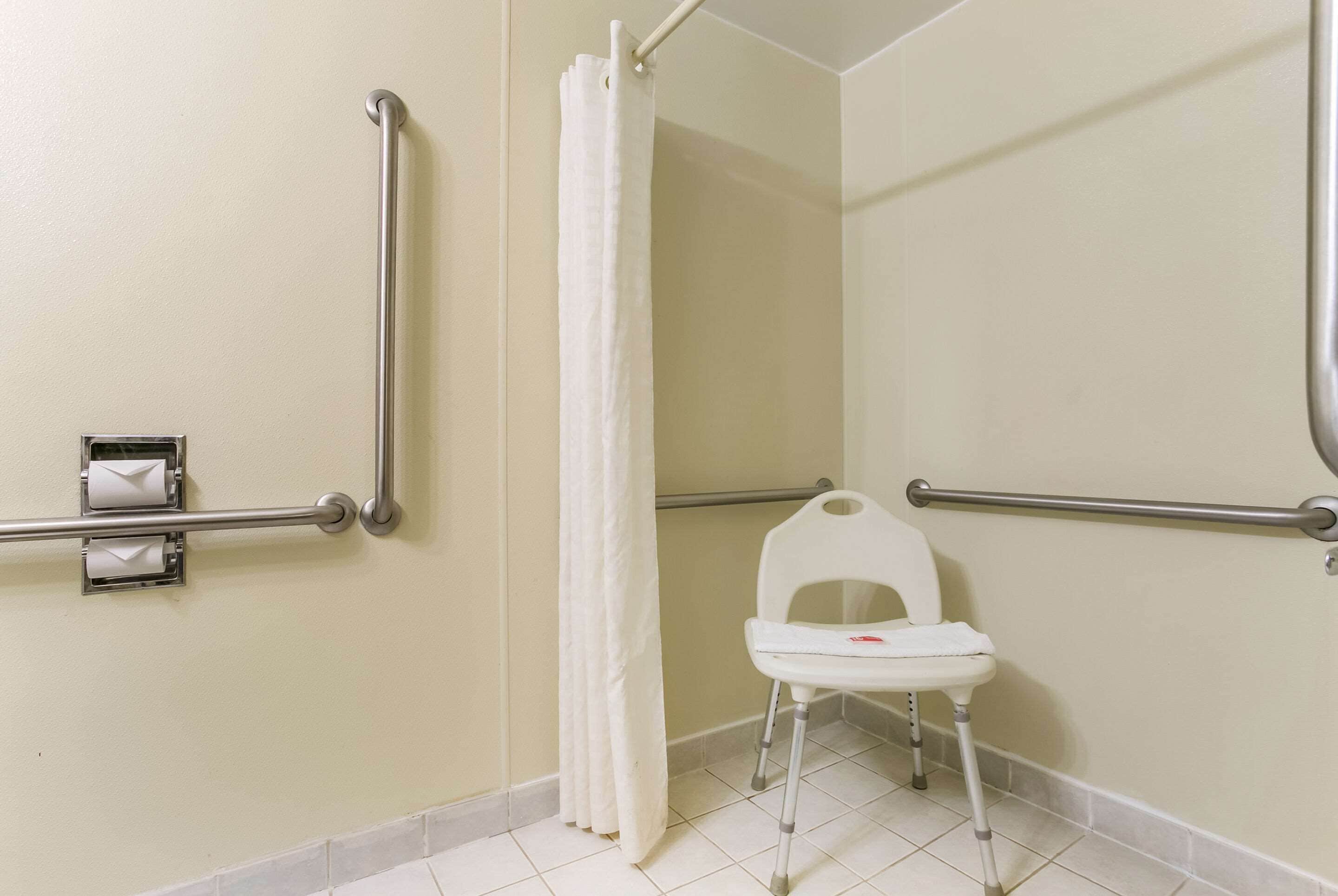 Uma casa de banho acessível em cadeira de rodas, com barras de apoio, um ecrã de chuveiro branco e uma cadeira branca.