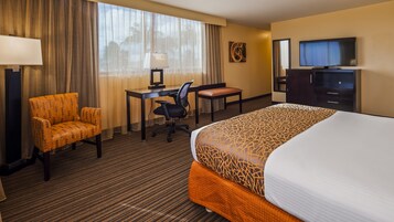 Quarto de hotel com uma cama grande, uma escrivaninha com computador, uma cadeira, uma televisão e um abajur.