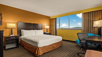 Quarto de hotel com uma cama grande, duas mesas de cabeceira com luminárias, uma escrivaninha com cadeira e vista para o exterior através de uma janela ampla.