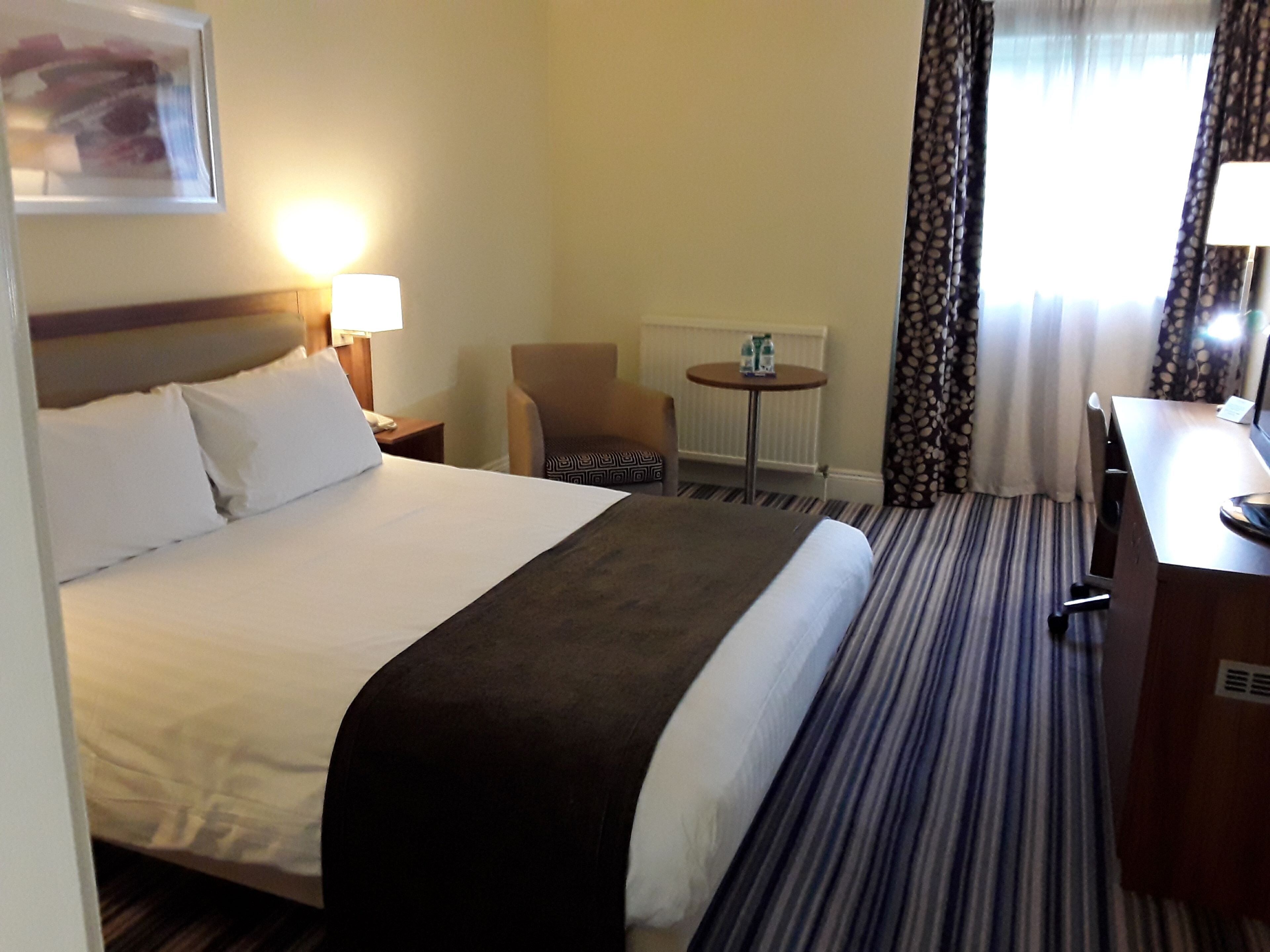Quarto de hotel com uma cama grande, uma mesinha com uma cadeira, uma escrivaninha com cadeira e uma janela com cortinas.
