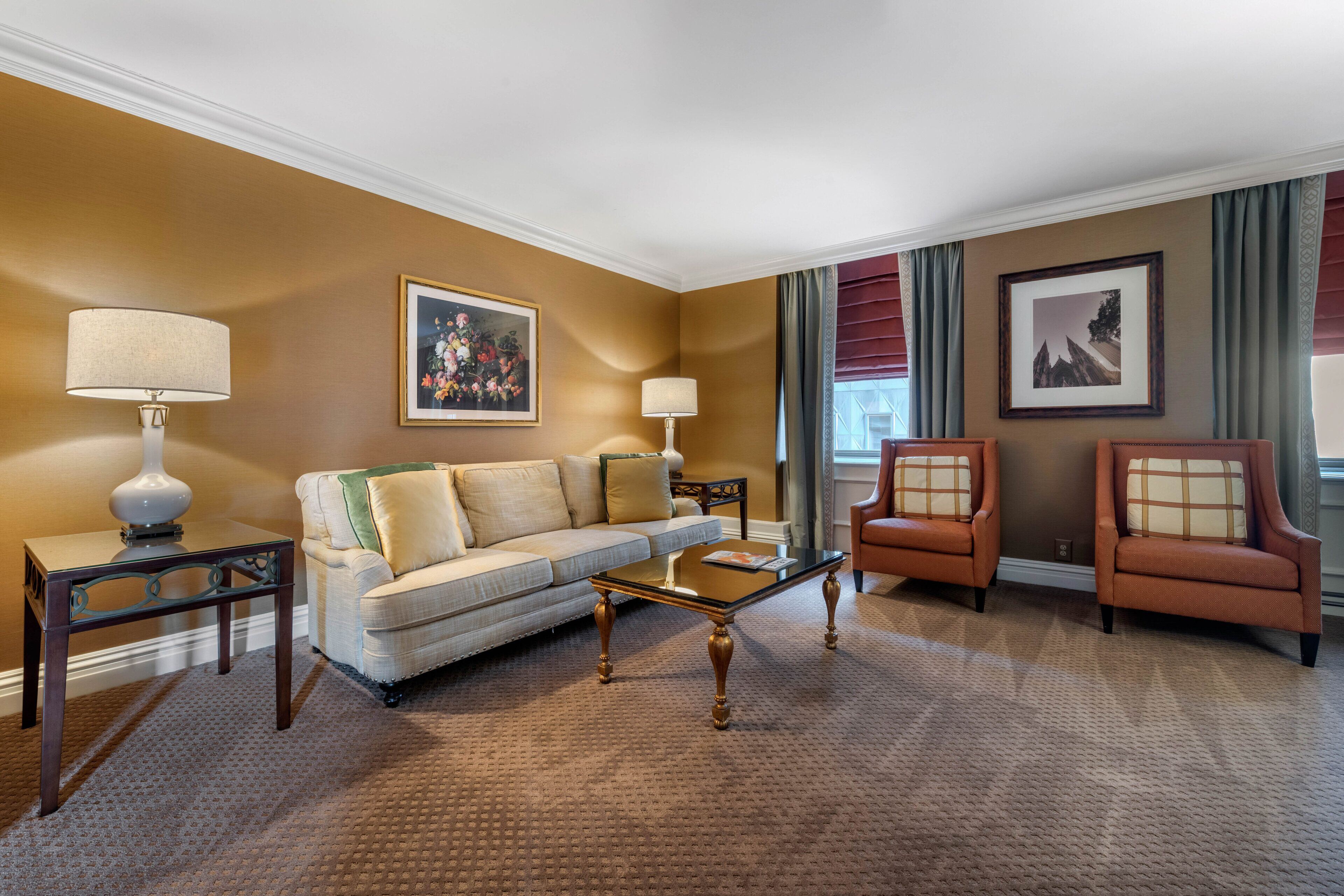Quarto de hotel com sofá bege, mesa de centro de vidro, mesa lateral com abajur e duas poltronas.
